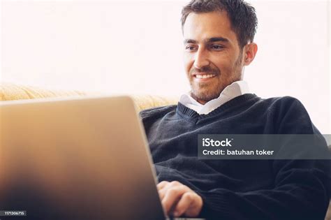따뜻한 친절한 미소로 카메라를 보고 소파의 팔에 균형 잡힌 노트북 컴퓨터와 함께 집에서 휴식 잘생긴 젊은 남자 35 39세에 대한 스톡 사진 및 기타 이미지 Istock