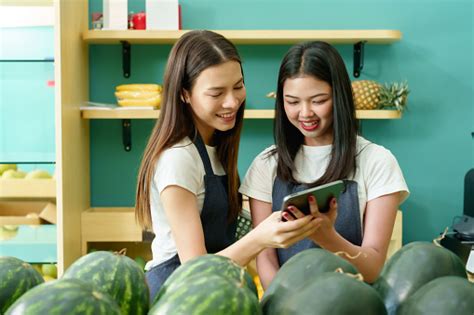 아시아 성인 여성과 백인 성인 여성이 과일 가게 안의 수박 선반 뒤에서 함께 태블릿을 보며 웃고 있는 모습은 Lgbtq 소상공인 커플의 팀워크와 기쁨과 연결을 반영합니다