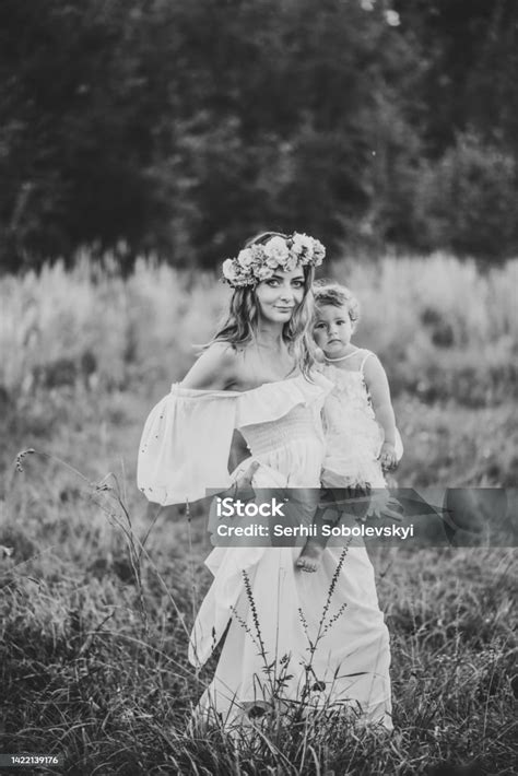 머리에 꽃 화환을 입은 딸을 입은 아름다운 젊은 어머니가 일몰 때 봄 밭에 서 있습니다 하루를 즐기는 아이가있는 엄마 여름 가족 사진 흑백 사진 2 3 살에 대한 스톡 사진