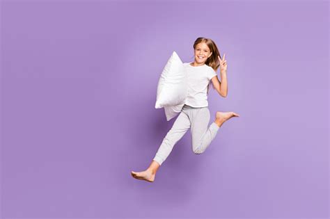 긍정적 인 쾌활한 귀여운 금발 머리 아이 점프 홀드 베개 를 즐길 수 주말 수면 파티 를 즐길 V사인 보라색 보라색 색상 배경 위에 고립 된 흰색 티셔츠 바지를 착용의 전체