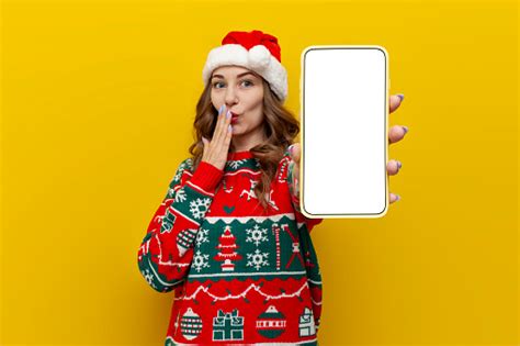크리스마스 스웨터와 산타 클로스 모자를 쓴 충격받은 소녀는 노란색 고립 된 배경에 스마트 폰의 빈 화면을 보여줍니다 12월에 대한 스톡 사진 및 기타 이미지 Istock