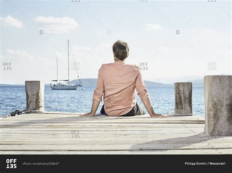 Back view of man sitting on jetty looking at distance stock photo - OFFSET