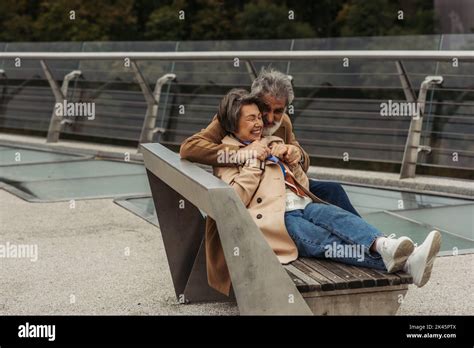 Happy Senior Man Hugging Positive Wife While Sitting On Bench Near Bridge Guard Rail Stock Image