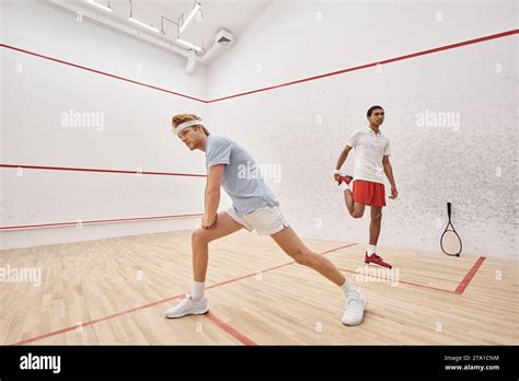 Sporty Redhead Man With Headband Doing Lunges Near African American Friend Inside Squash Court