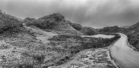 Greyscale Of Hills Covered In The Grass Surrounded By A Road And A Lake
