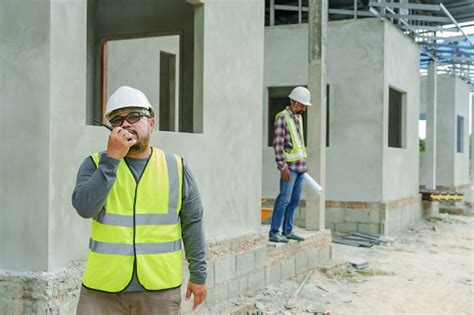 건설 안전 조끼를 입은 남성이 전화 통화를 하고 있고 안전모와 청사진을 쓴 또 다른 남성이 미완성 건물 계단에 서 있다 건축가와