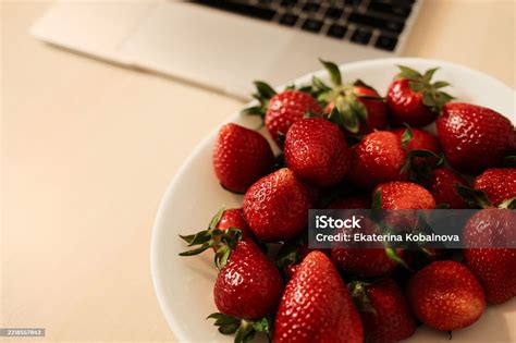 잘 익은 빨간 딸기가 가득한 하얀 그릇이 밝은 나무 책상 위의 열린 은색 노트북 옆에 놓여 있으며 깨끗하고 매력적인 작업 공간 환경에서 생생한 과일과 현대 기술이 조화를