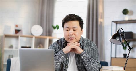 체크 무늬 셔츠를 입은 사려 깊은 아시아 남자가 집에서 테이블에 앉아 컴퓨터 화면을 보고 있다 젊은 프리랜서는 아이디어를 고려하거나 작업 순간에 대한 결정을 내립니다