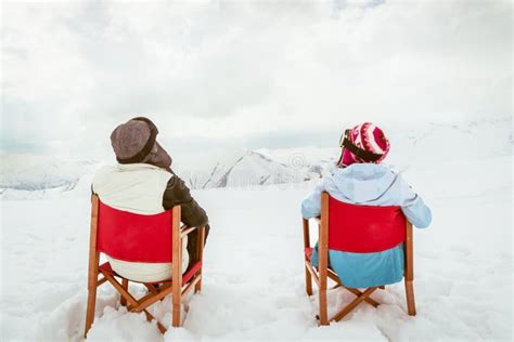 Back View Couple Sitting In Deck Chairs By Ski Slope Enjoying Snowy