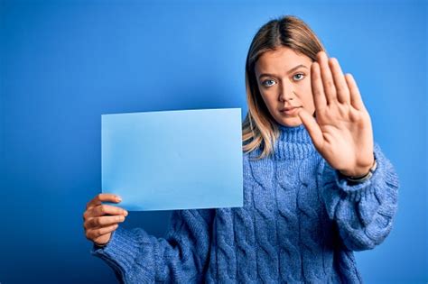 진지하고 자신감있는 표정 방어 제스처로 정지 표지판을하고 열린 손으로 고립 된 파란색 배경 위에 서있는 배너를 들고있는 젊은