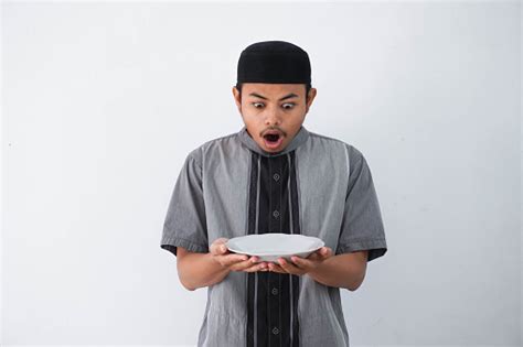 하얀 배경에 고립된 회색 무슬림 옷을 입고 빈 저녁 식사 접시를 들고 흥분한 표정을 짓고 있는 젊은 아시아 무슬림 남성이 충격을