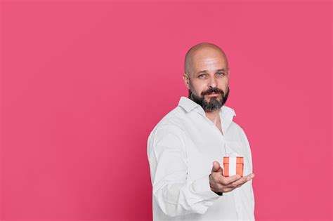 흰색 셔츠를 입은 잘생긴 진지한 중년 남성이 분홍색 배경에 미소 짓는 흰색 활로 장식 된 빨간 선물을 손에 들고 있습니다 40 49세에 대한 스톡 사진 및 기타 이미지