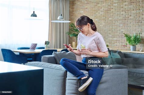 집 소파에 앉아 스마트폰을 사용하는 물 한 잔을 들고 있는 중년 여성 40 49세에 대한 스톡 사진 및 기타 이미지 40 49세 45 49세 Brand Name