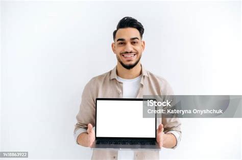 즐거운 잘 생긴 인도 또는 아라비아 청년 학생 또는 프리랜서 흰색 고립 된 배경에 서서 프리젠 테이션을 위해 빈 흰색 모형 화면이있는 열린 노트북을 들고 카메라를보고 미소