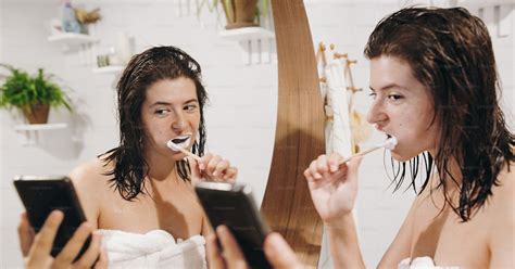 소셜 미디어가 영향을 미칩니다 하얀 수건을 쓴 젊은 행복한 여자가 이를 닦고 둥근 거울에 있는 세련된 욕실에서 전화 화면을