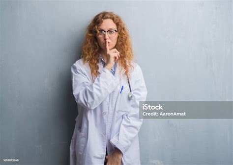 입술에 손가락으로 조용히 하 라는 코트를 입고 회색 그런 지 벽에 빨강 머리 젊은 의사 여자 침묵 그리고 비밀 개념입니다 건강