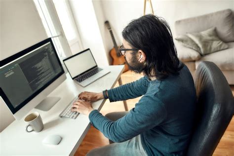 Young Male Web Developer Working From Home Using Desktop Computer And Laptop Writing Code