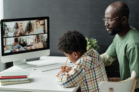 어린 소년은 선생님과 함께 책상에 앉아 글을 쓰고 온라인 대화 중에 컴퓨터로 공부하고 있습니다 사진 Unsplash의 흑인 소년 이미지
