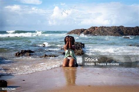 검은 파워 머리를 가진 아름다운 여자의 초상화는 해변 모래에 무릎을 꿇고 물을 던진다 푸른 하늘 바위와 구름을 배경으로 한다