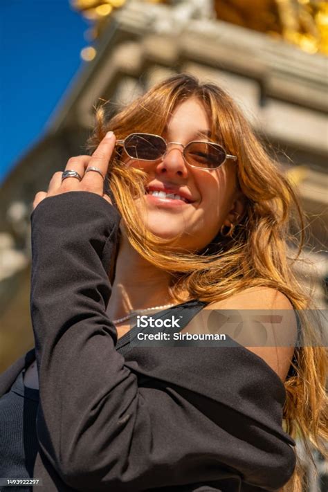 그녀의 긴 금발 머리를 만지고 선글라스와 검은 색 탑을 입고 사랑스럽고 아름다운 햇볕에 쬐인 얼굴과 어깨에서 떨어지는 검은 색 재킷으로 행복하게 웃고 웃는 화려한 젊은 모델