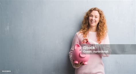 서 치아를 보여주는 자신감 미소로 웃 고 행복 한 얼굴로 저금통 달러를 들고 회색 그런 지 벽에 빨강 머리 젊은 여자 저축에 대한 스톡 사진 및 기타 이미지 Istock