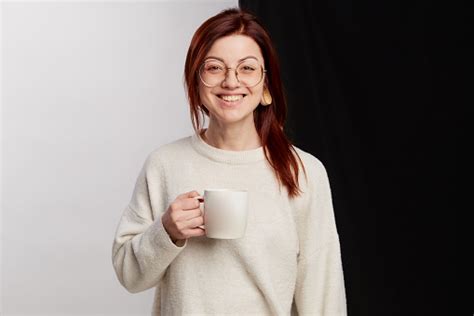 뜨거운 음료가 활기를 띠고 있습니다 차 또는 커피 한 잔을 손에 들고 있는 빨간 머리 여자 큰 안경에 반쯤 흰색과 검은 색