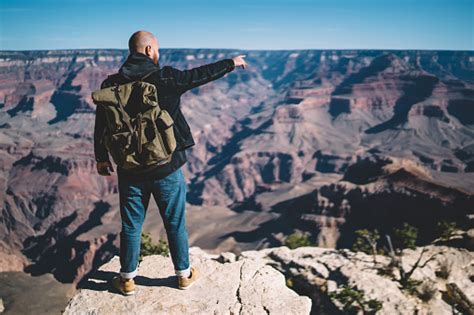 바위 높은 봉우리에 서있는 하이킹 루트를 가리키는 배낭을 가진 남성 여행자의 뒷모습 그랜드 캐년 국립 공원의 풍경 풍경에서 멀리 떨어진 무언가를 보여주는 힙스터 남자 방랑자