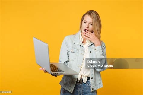 스튜디오에서 노란색 오렌지 벽 배경에 고립 된 노트북 Pc 컴퓨터를 사용 하 여 손으로 들고 입을 덮고 데님 캐주얼 의류에 충격 젊은 여자 사람들 라이프 스타일 개념입니다