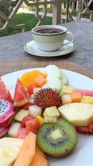 생생하게 자른 신선한 키위 드래곤 과일 수박 조각이 하얀 접시에 담겨 있으며 햇살이 내리쬐는 야외 환경에서 김이 모락모락 나는 아침 음료와 함께 제공됩니다 0명에 대한 스톡