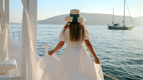 긴 머리에 모자를 쓰고 긴 흰색 드레스를 입은 젊은 갈색 머리 여성이 석양이 비추는 바다의 나무 부두에서 달리고 있습니다 관광 여름철 휴가 및 해변에서의 휴가 해변에 대한