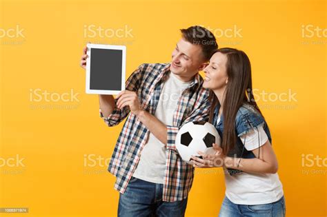 젊은 커플 팬 여자 사람 지원 팀을 응원 하는 축구 팬 들 재미 있는 노란색 배경에 고립 된 축구 공을 빈 검은색 빈 화면으로 태블릿 Pc 컴퓨터를 들고 스포츠 가족 라이프