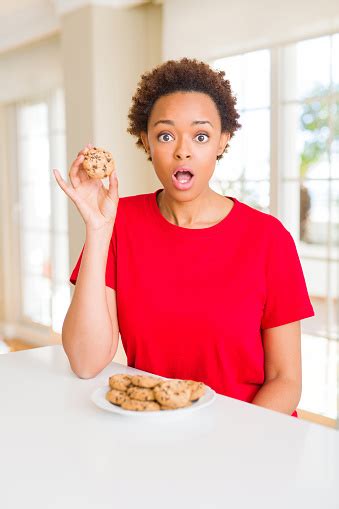 초콜릿 칩 쿠키를 먹는 젊은 아프리카 계 미국인 여성은 깜짝 얼굴로 충격에 무서워하고 두려움과 두려움 표현으로 흥분했습니다 감정에 대한 스톡 사진 및 기타 이미지 Istock