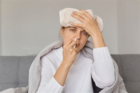 이마에 젖은 수건을 든 아픈 화가 난 여성의 초상화가 소파에 앉아 비강 스프레이를 사용하여 콧물 살포를 사용하여 고온을 가진 담요에 싸여 있습니다 거실에 대한 스톡 사진 및