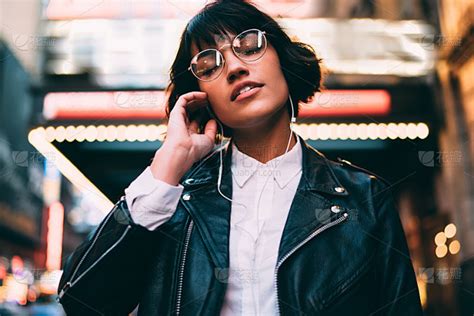 黑发时髦的时髦女孩，戴着光学眼镜，戴着耳机听电子音乐，在自由的时间里，站在纽约市中心的夜晚，在昏暗的