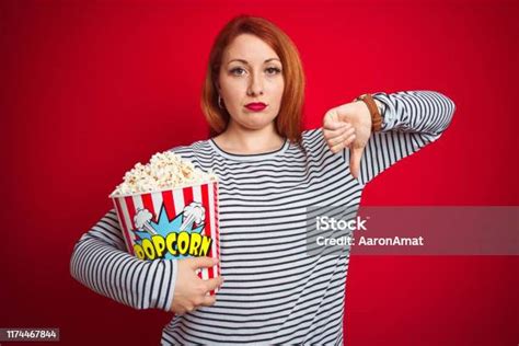 화가 얼굴 엄지 손가락으로 싫어하는 것을 보여주는 부정적인 기호 거부 개념으로 빨간 고립 된 배경 위에 팝콘을 먹는 젊은 아름다운 빨간 머리 여자 T 셔츠에 대한 스톡 사진