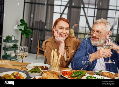 Happy Redhead Woman Smiling Near Bearded Husband With Wine Glass Near Delicious Supper In Living
