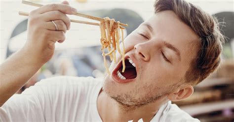 세련된 배고픈 남자는 대나무 젓가락으로 판지 상자에서 야채와 함께 맛있는 냄비 국수를 먹고 있습니다 아시아 거리 음식 축제