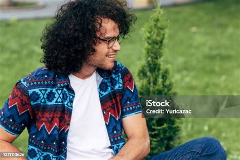 곱슬 머리를 웃고 화려한 셔츠와 공원벤치에 앉아 투명 안경을 입고 백인 젊은이 화창한 여름날을 즐기는 캐주얼 한 트렌디 한 의상을 입은 쾌활한 남성 20 29세에 대한 스톡