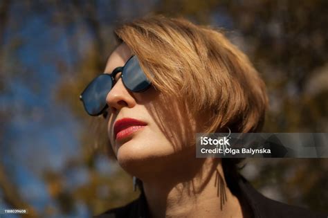 짧은 머리와 거울 표면 선글라스를 가진 젊고 아름다운 소녀는 푸른 하늘과 노란 잎 배경이가있는 나무를 배경으로 가을 공원에서 시간을 보내고 있습니다 가을에 대한 스톡 사진