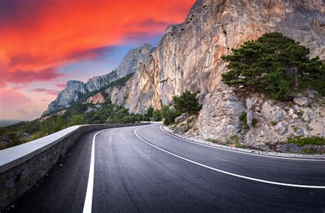 아스팔트도로 완벽 한 아스팔트 높은 바위 나무 여름의 석양에 붉은 구름이 있는 놀라운 하늘을 가진 아름 다운 권선 산악 도로가 있는 풍경 파노라마 여행 배경입니다 산악 고속