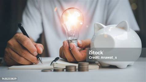에너지 절약과 돈 개념 저축 또는 투자를위한 아이디어 돼지 저금통에 전구를 들고 돈 금융 계획을위한 메모가있는 책상에 쌓인 동전을 들고있는 사업가 저축에 대한 스톡 사진 및