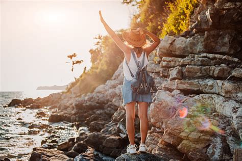 모자를 쓴 젊은 힙스터 여성과 손을 들고 루크작을 들고 절벽 꼭대기에 서서 해질녘에 바다를 바라 보았다 20 24세에 대한 스톡 사진 및 기타 이미지 Istock