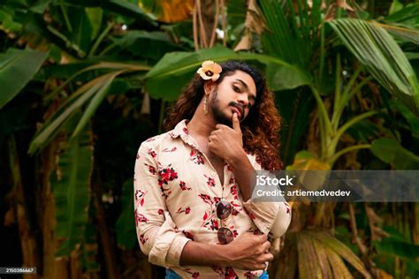 머리에 꽃을 꽂은 스리랑카 게이 남성이 열대 지방에 서서 턱을 만지며 생각에 잠긴다 그의 플로럴 셔츠는 풍부한 식물과 조화를 이루며 정체성과 부드러운 남성미를 표현합니다