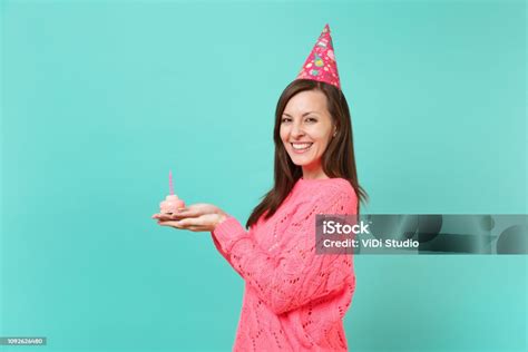 미소 니트 핑크 스웨터 생일 모자에 있는 젊은 여자의 측면 보기 손에 촛불 블루 청록색 배경 스튜디오 초상화에 고립 된 케이크 개최 사람들의 라이프 스타일 개념입니다 복사