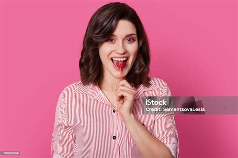 아름 다운 여성 모델의 사진 입에 사탕을 보유 하 고 여가 시간을 즐기고 줄무늬 셔츠를 입고 카메라에서 직접 분홍색 배경 감싸고 달콤한 사탕에 포즈 핀 업 소녀는 미소 및