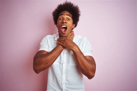 고립 된 분홍색 배경 위에 서있는 흰색 셔츠를 입은 아프리카 머리를 가진 젊은 미국 남자는 고통스러운 목졸림 때문에 외치고 질식합니다 건강 문제 질식 과 자살 개념 건강