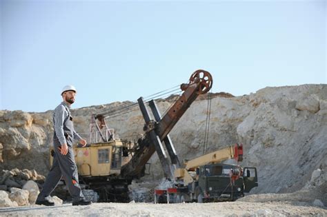 Premium Photo A Worker In A Helmet Stands On The Background Of An Excavator In A Quarry