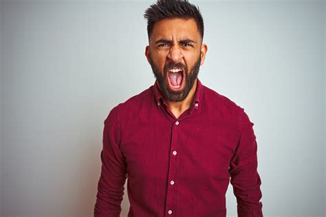 고립 된 회색 배경 위에 서있는 빨간 우아한 셔츠를 입은 젊은 인도 남자는 분노와 분노로 외치며 좌절하고 분노한 비명을 지르고 있습니다 분노와 공격적인 개념 감정에 대한