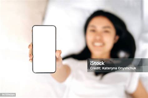 광고 또는 프리젠 테이션을위한 모형 개념 집중이 잘되지 않은 아시아 소녀는 침대에 누워 웃고 빈 흰색 화면이있는 스마트 폰을