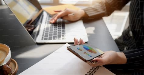 편안한 직장에서 다른 사무용품과 함께 스마트폰으로 비즈니스 차트를 보면서 노트북으로 작업하는 사업가의 측면 모습 사진